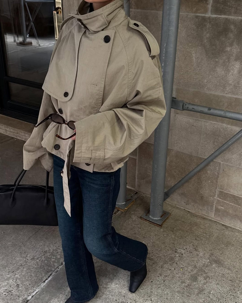 Stylish woman wearing a beige oversized trench coat%2C dark blue jeans%2C and black pointed-toe boots%2C holding sunglasses and a black leather tote bag%2C standing on a city sidewalk.
