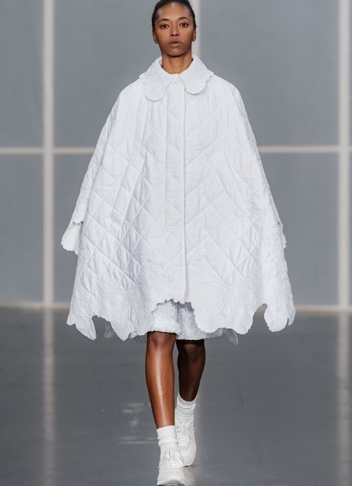 Cecilie Bahnsen FW20 collection showcases a model in a striking white quilted cape with scalloped edges, layered over a delicate dress and paired with sporty white sneakers