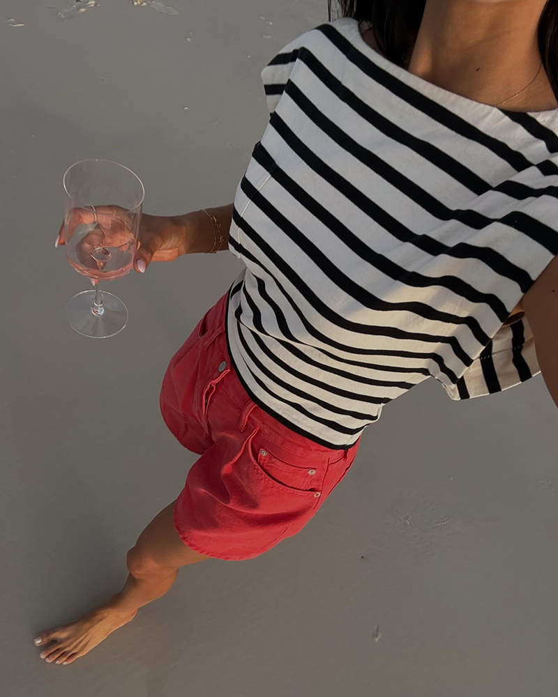 Close-up of striped black-and-white knit top with red shorts and wine glass%2C styled barefoot on sandy beach at sunset