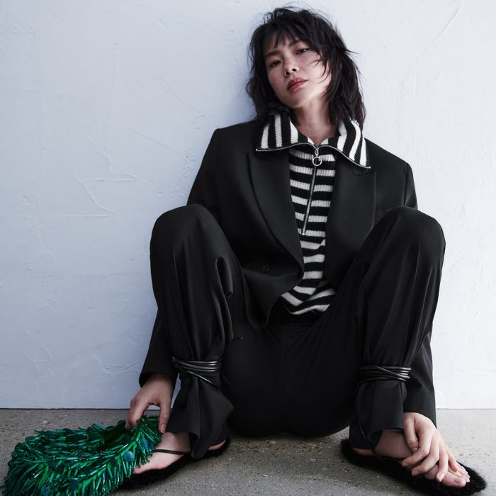 A woman sitting on the floor wearing a striped sweater under a black suit.