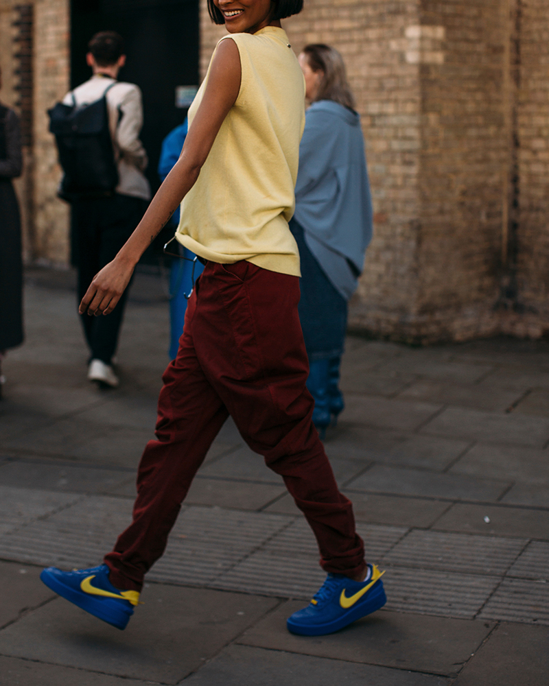 Street style look featuring a yellow sleeveless knit%2C burgundy trousers and blue Nike sneakers on a city sidewalk