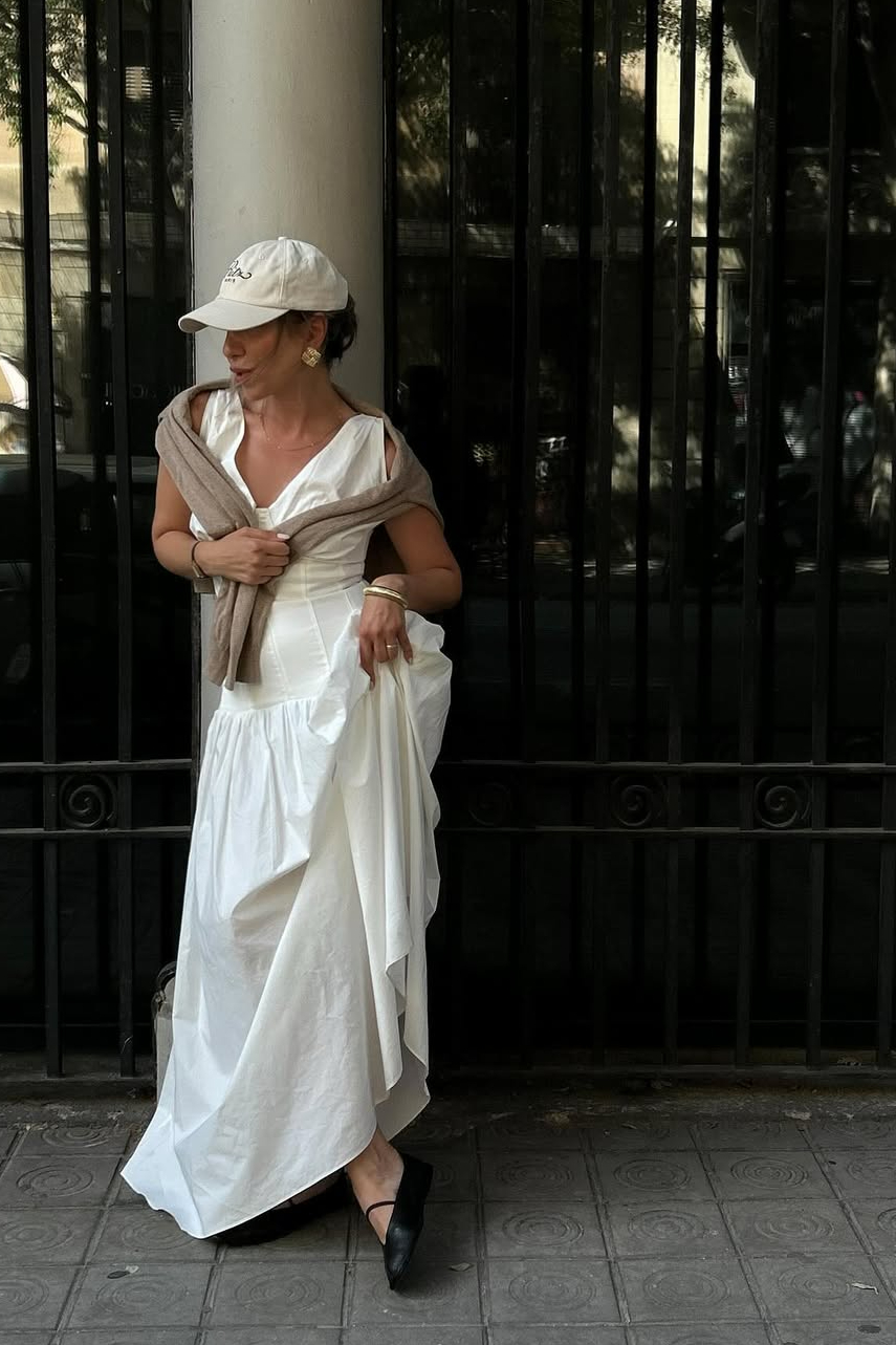 White midi dress with a flared hemline%2C styled with a beige knit wrap and a white cap%2C complemented by black flats%2C exuding a chic neutral look