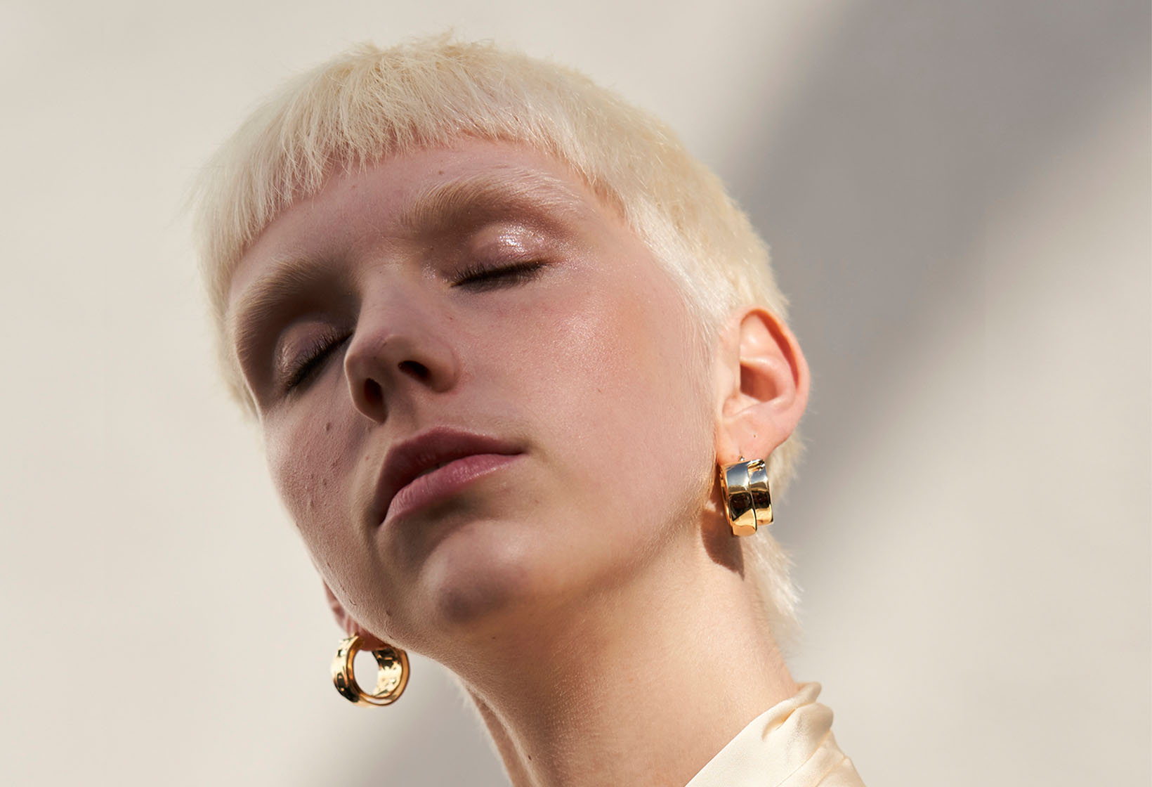 Minimalist style portrait featuring a model with platinum blonde hair and gold hoop earrings%2C showcasing a natural makeup look against a soft backdrop