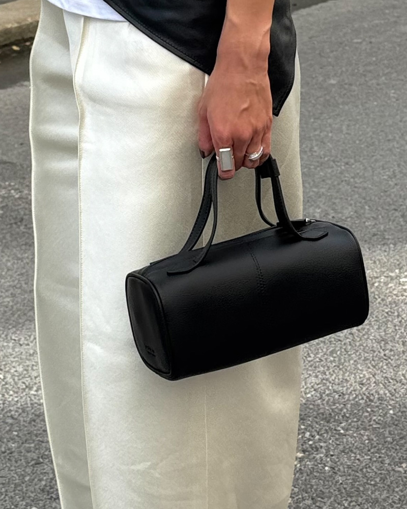 Close-up of black leather barrel bag with top handles%2C styled with ivory satin skirt and minimal jewelry on city street