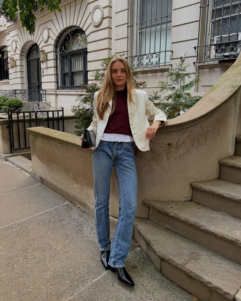 Model in a white blazer%2C burgundy top and straight-leg blue jeans with black boots on a city sidewalk