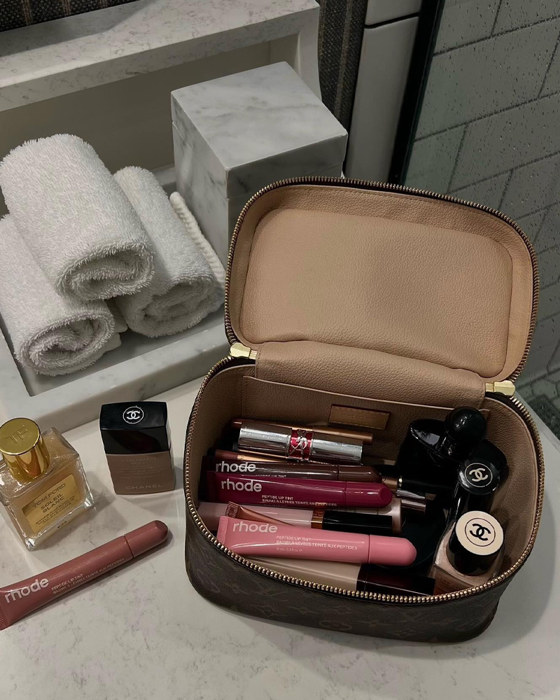 Louis Vuitton monogram toiletry bag filled with Rhode lip tints%2C Chanel makeup and Tom Ford perfume on a marble bathroom counter