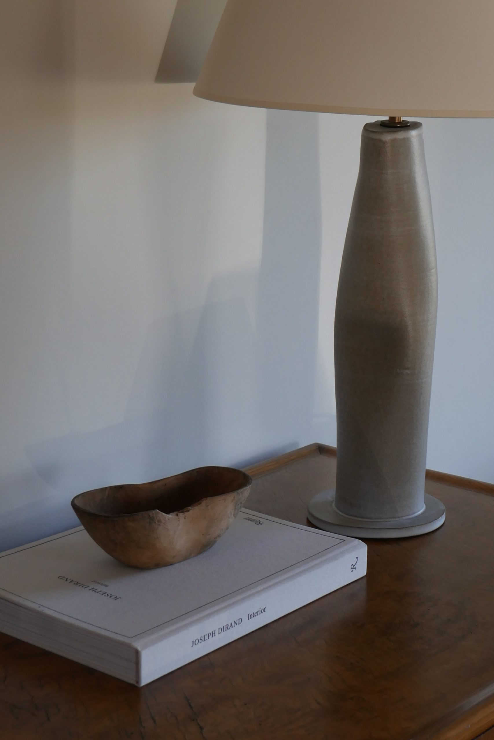 Minimalist decor featuring a sleek gray ceramic lamp with a beige shade%2C complemented by a wooden bowl atop a coffee table book titled %22Joseph Dirand Interior%22
