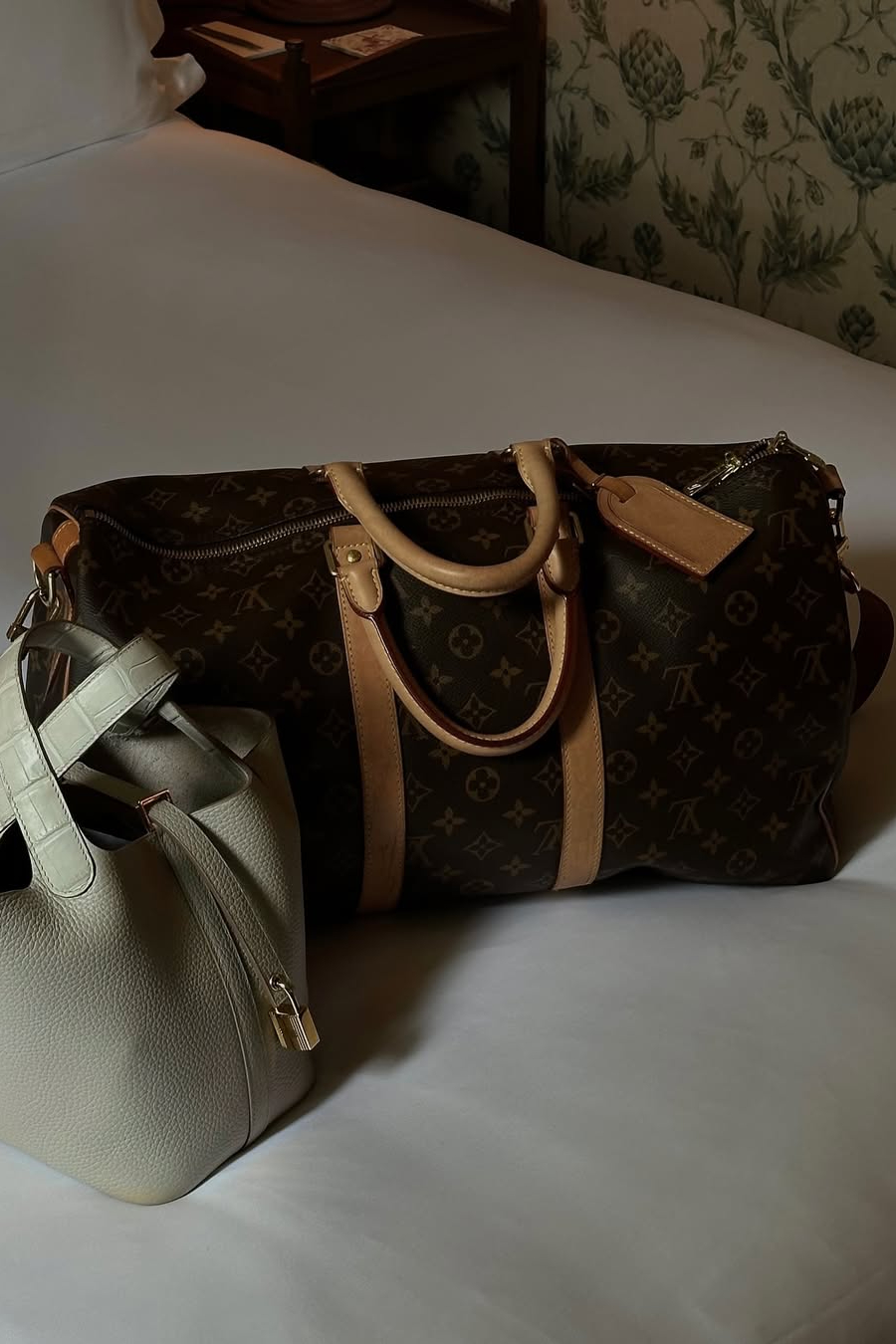 Louis Vuitton Keepall bag in classic monogram canvas with tan leather handles%2C paired with a soft gray leather handbag%2C styled on a white bed