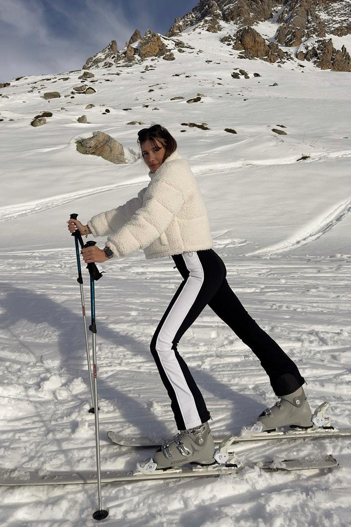 Cozy cream puffer jacket paired with sleek black ski pants featuring white stripes%2C set against a snowy mountain backdrop