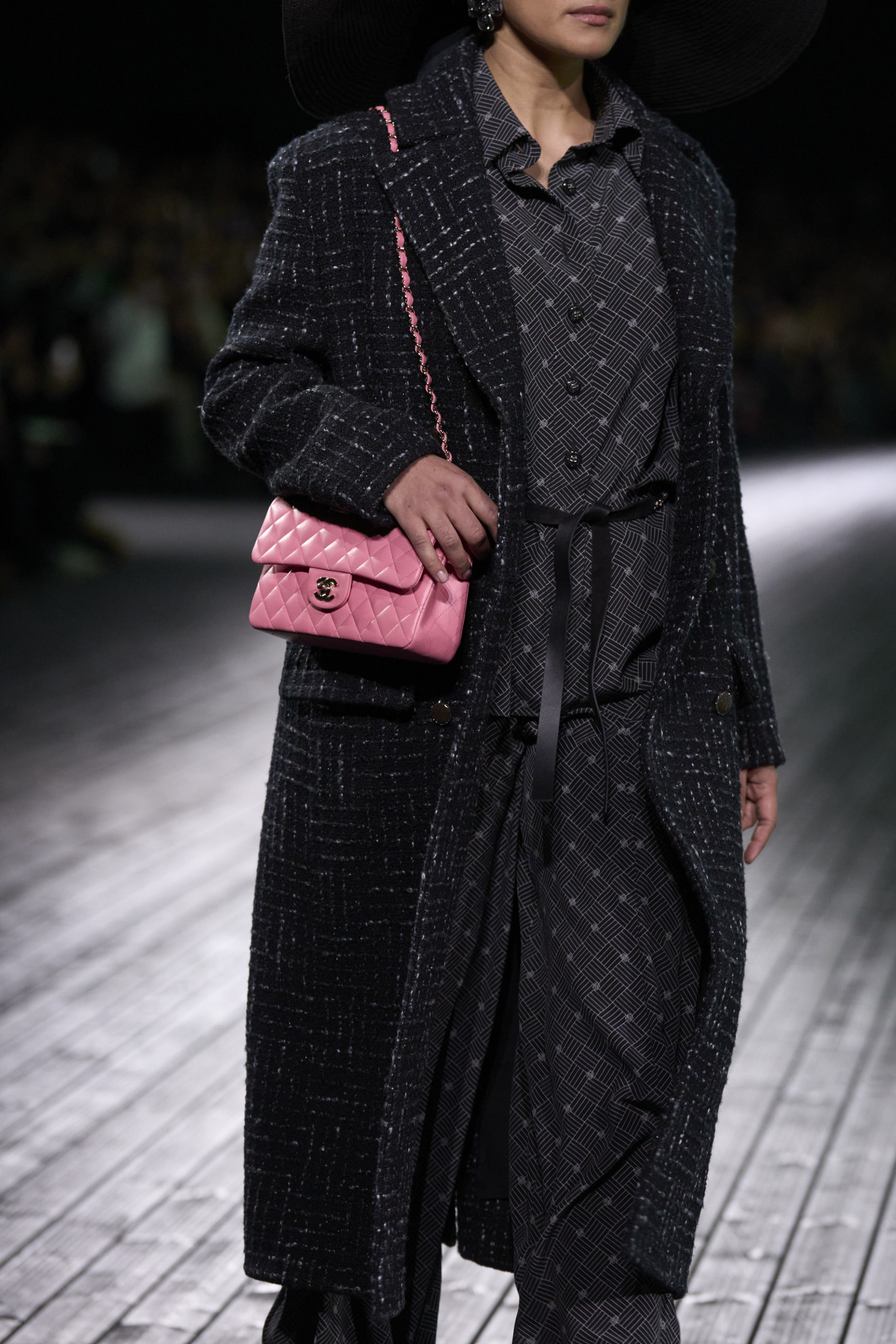 Model wearing a black textured Chanel coat over a patterned black jumpsuit, accessorized with a pink quilted Chanel flap bag with chain strap