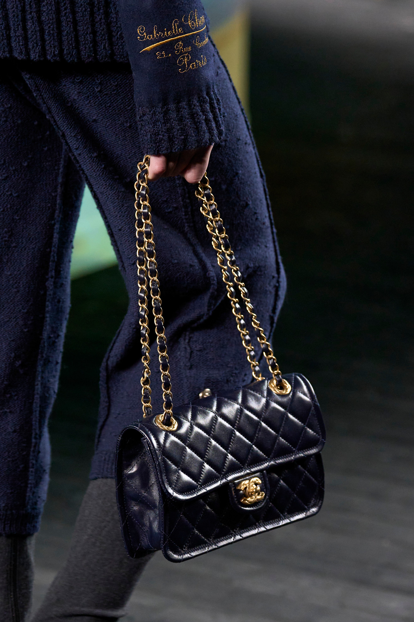 Quilted black leather Chanel classic flap bag with gold chain straps held by model in navy knit outfit