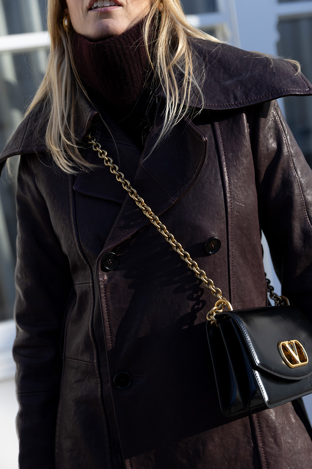 Valentino crossbody bag in luxurious black leather%2C featuring a gold logo clasp%2C elegantly paired with a stylish oversized brown coat