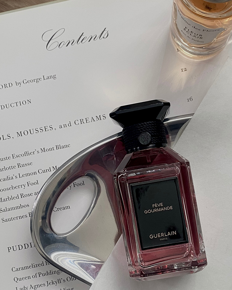Guerlain F%C3%A8ve Gourmande perfume bottle with deep burgundy glass and black label%2C styled on an open book with a silver spoon and soft tabletop setting