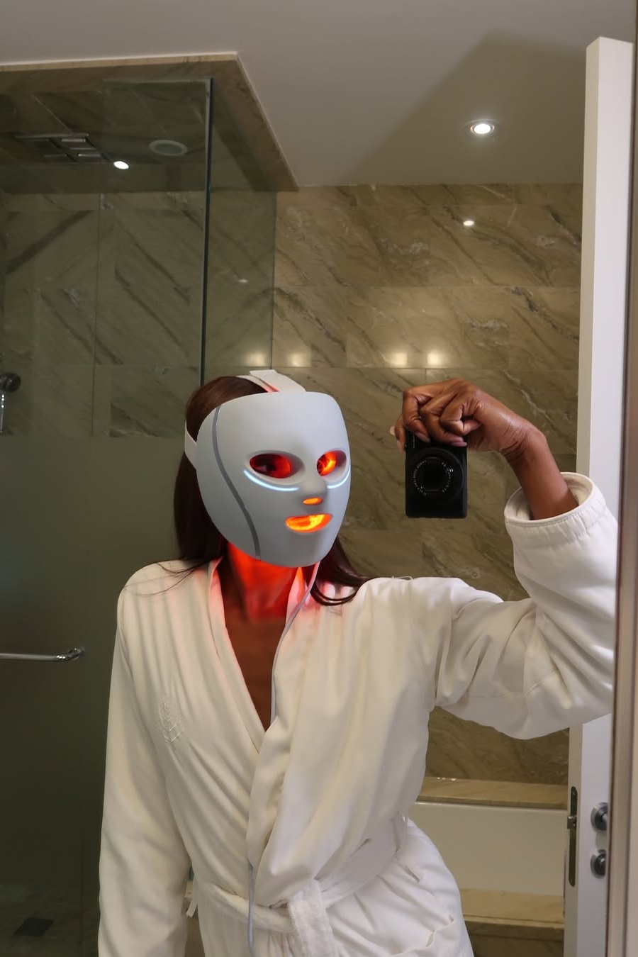 High-tech red light therapy mask by a luxury brand%2C worn by a woman in a plush white bathrobe%2C showcasing a spa-like setting