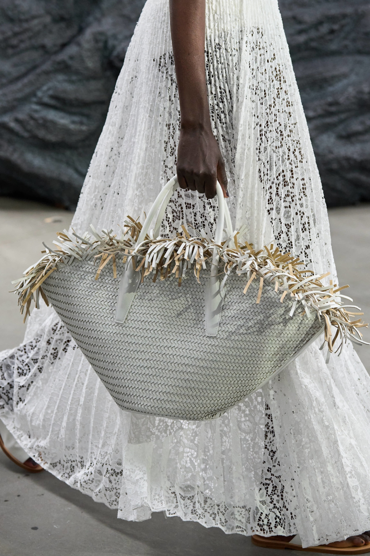 Woven summer tote bag with textured surface and decorative fringe detail%2C paired with a flowing white lace dress for a chic look