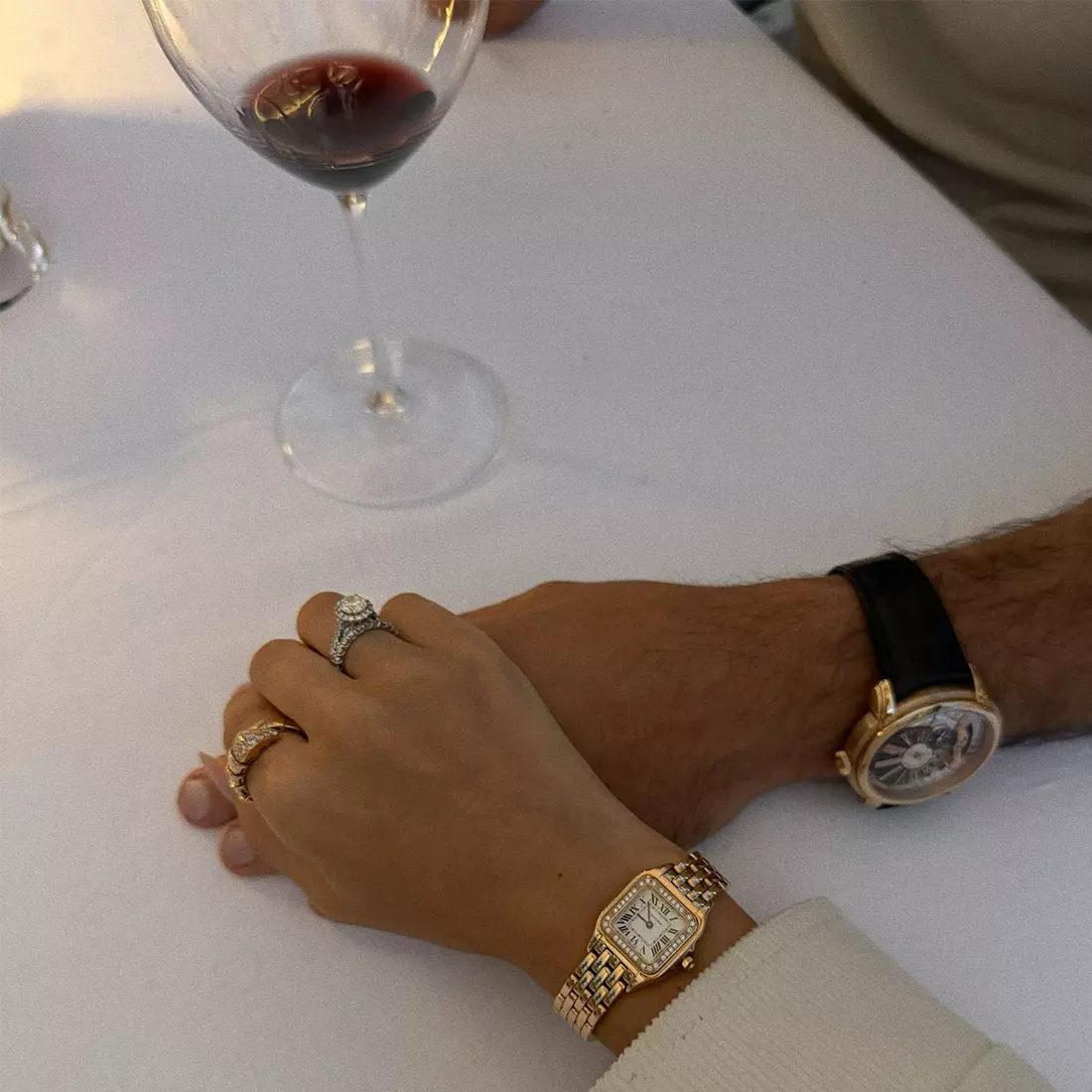 Elegant couple%27s watch display featuring a gold-tone square watch with a silver bracelet on a woman%27s wrist%2C alongside a classic black leather watch on a man%27s wrist%2C accentuated by sparkling rings and a refined dining setting