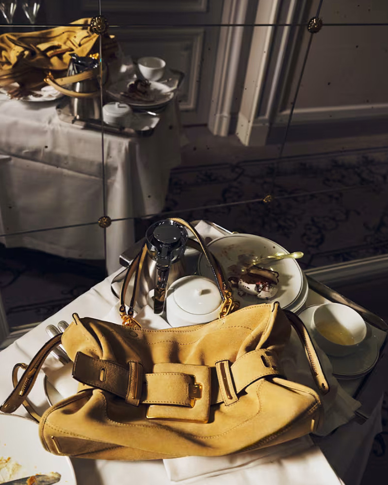 Balmain suede shoulder bag with oversized buckle hardware in camel leather%2C styled on a caf%C3%A9 table with porcelain cups and mirrored backdrop