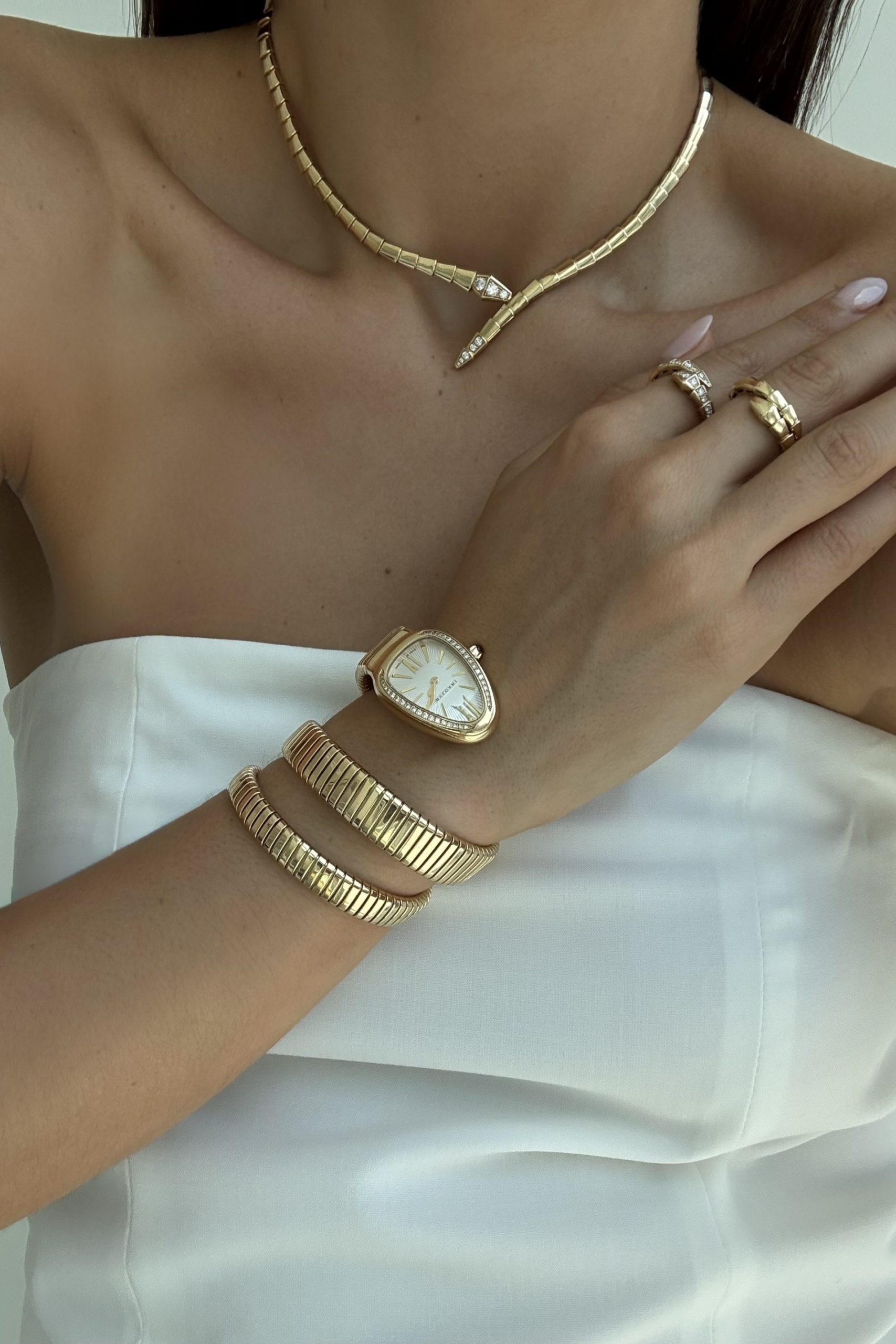 Gold snake chain necklace paired with a classic gold watch%2C stackable bangles%2C and elegant rings against a chic white top