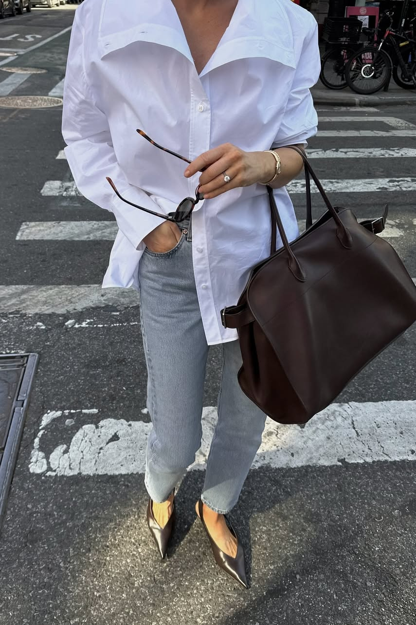 Chic woman in a crisp white oversized shirt styled with light blue high-waisted jeans and black pointed-toe mules%2C accessorized with a dark leather handbag and sunglasses