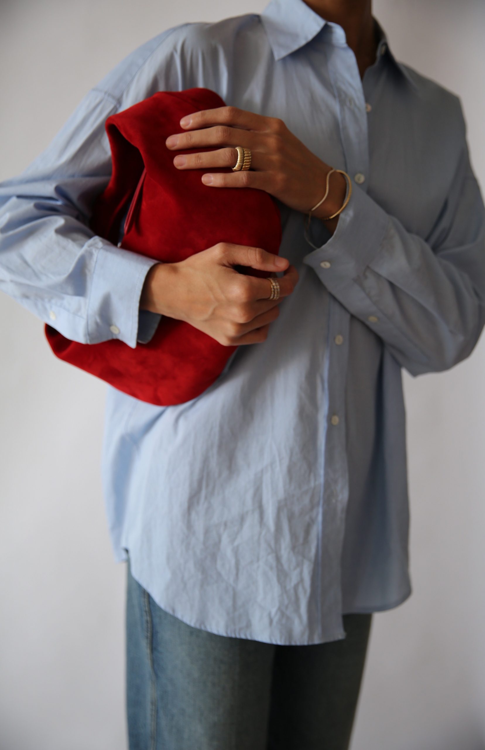 Khaite Olivia hobo bag in vibrant red suede%2C elegantly held by a model dressed in a light blue oversized shirt and layered rings
