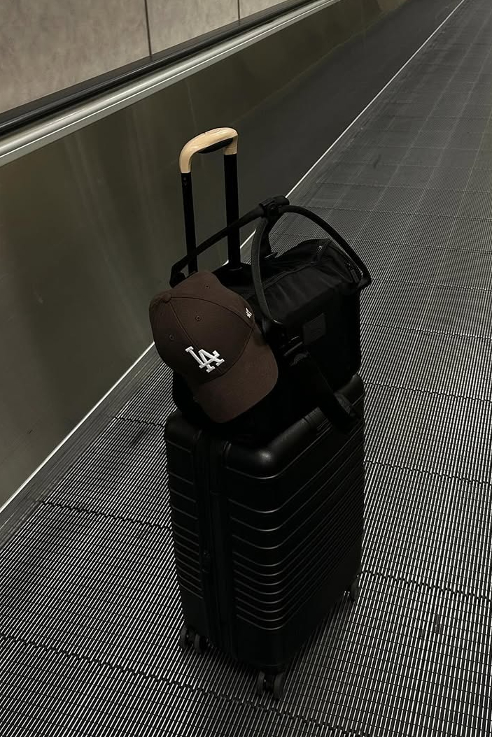 Black hard-shell carry-on luggage with a sleek design%2C topped with a brown LA Dodgers cap%2C set against a modern airport backdrop