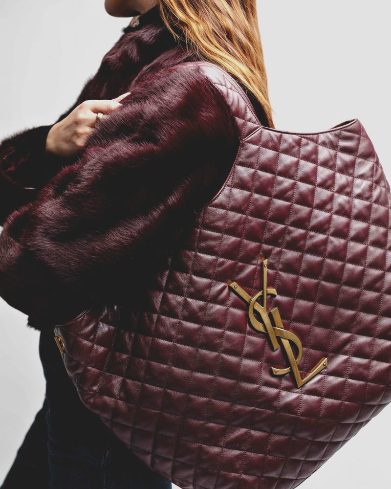 Saint Laurent quilted burgundy leather tote bag features a signature gold YSL emblem%2C elegantly paired with a luxurious faux fur jacket for a chic look
