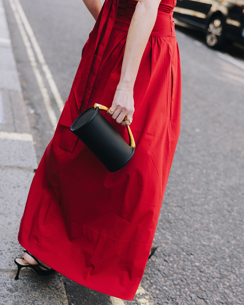 A woman in a vibrant red maxi dress carries a sleek black bucket bag with a gold handle%2C showcasing a chic urban look