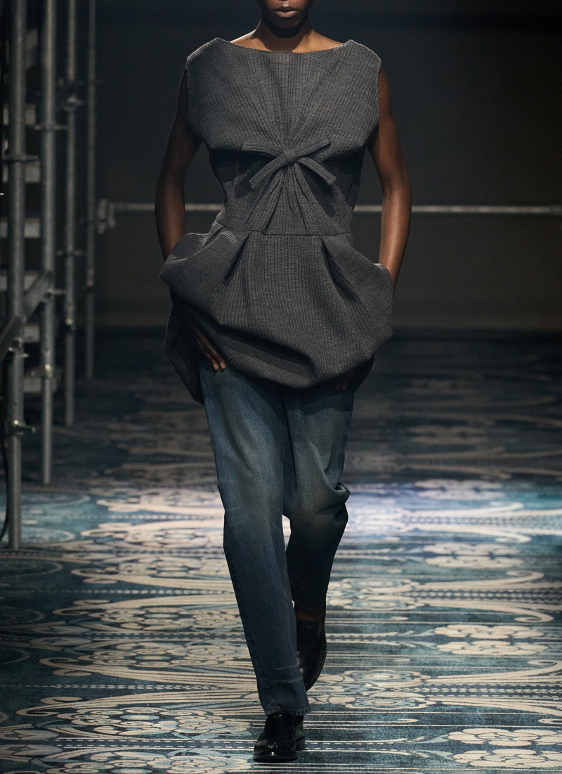 Model wearing structured gray textured top with bow detail and relaxed prada jeans on dimly lit runway