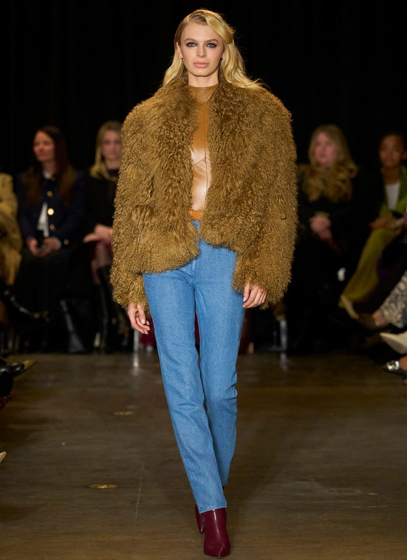 Model wearing tan shearling jacket with satin blouse, Sergio Hudson blue jeans, and burgundy boots on runway