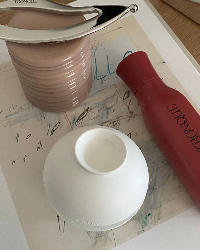 Tranquil beauty display featuring a sleek silver facial tool%2C organic-shaped pink storage pots%2C and a vibrant red hair product%2C arranged artfully on a textured backdrop
