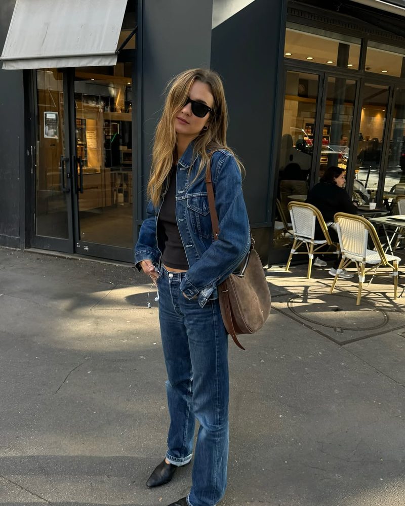 Casual chic outfit featuring a denim jacket over a black crop top%2C paired with high-waisted jeans and black leather mules%2C complemented by stylish sunglasses and a brown handbag.