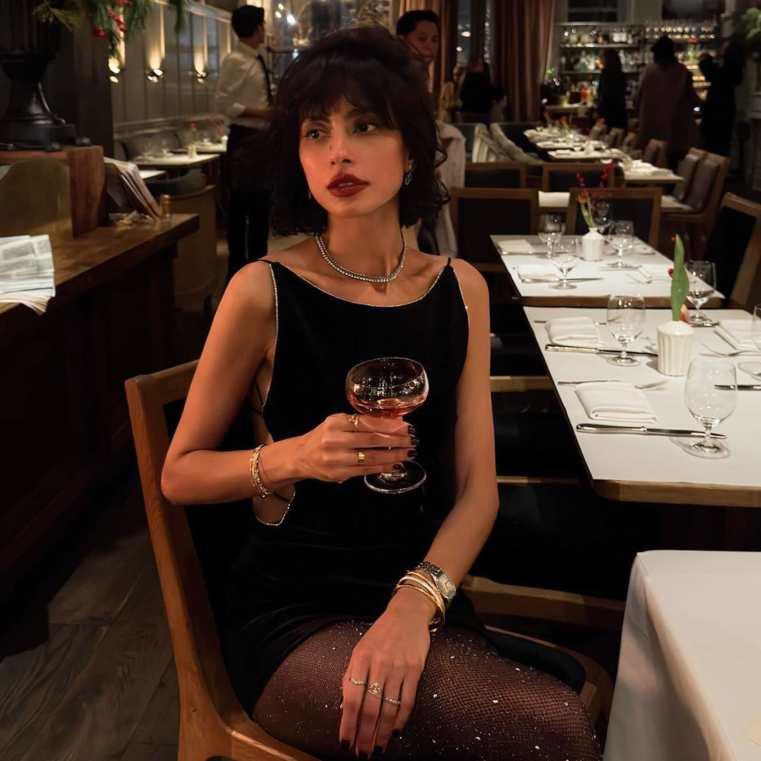 Elegant black velvet mini dress with sparkling details%2C paired with layered silver jewelry and a chic cocktail glass in a luxe dining setting