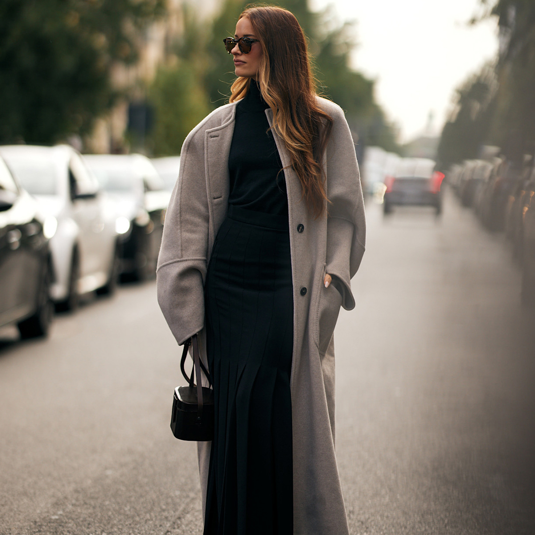 Chic winter outfit featuring a light grey oversized coat layered over a sleek black turtleneck and a long black skirt%2C accessorized with stylish sunglasses and a small handbag
