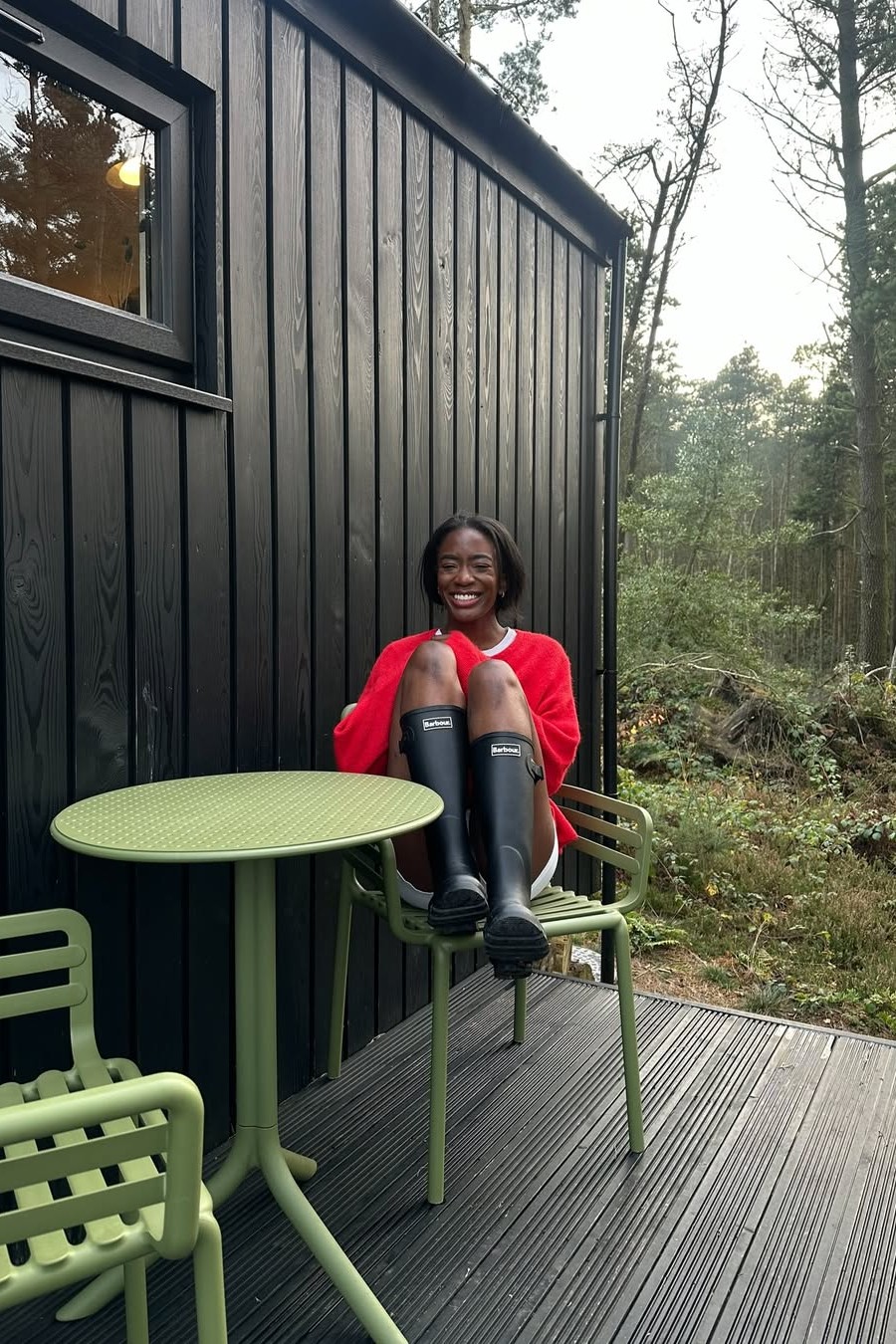 Outdoor cottage scene with Barbour black rain boots styled with a red sweater on a modern deck surrounded by trees