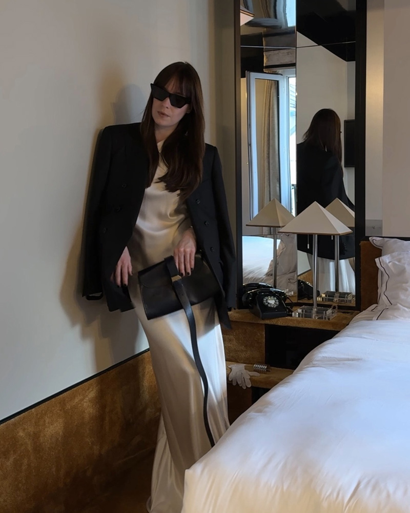 Woman in cream satin slip dress with black blazer%2C sunglasses%2C and leather clutch in a modern hotel room