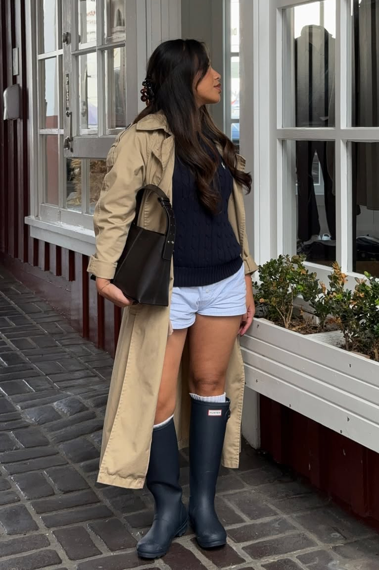 Stylish rain outfit featuring a beige trench coat%2C navy knit sweater%2C blue shorts%2C and sleek black Hunter rain boots%2C set against a charming urban backdrop