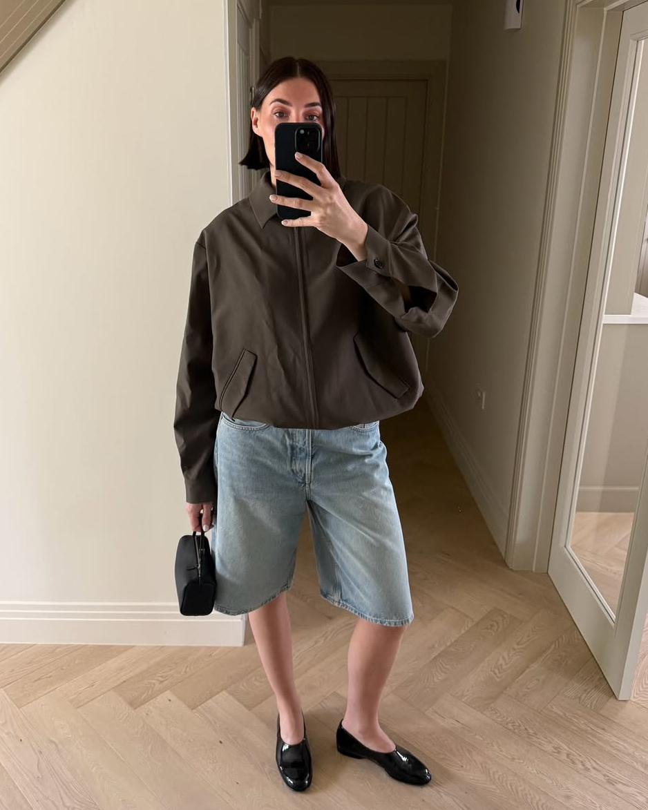 Mirror selfie showcasing a cropped brown utility jacket styled with light denim Bermuda shorts%2C black patent loafers%2C and a mini top-handle bag indoors