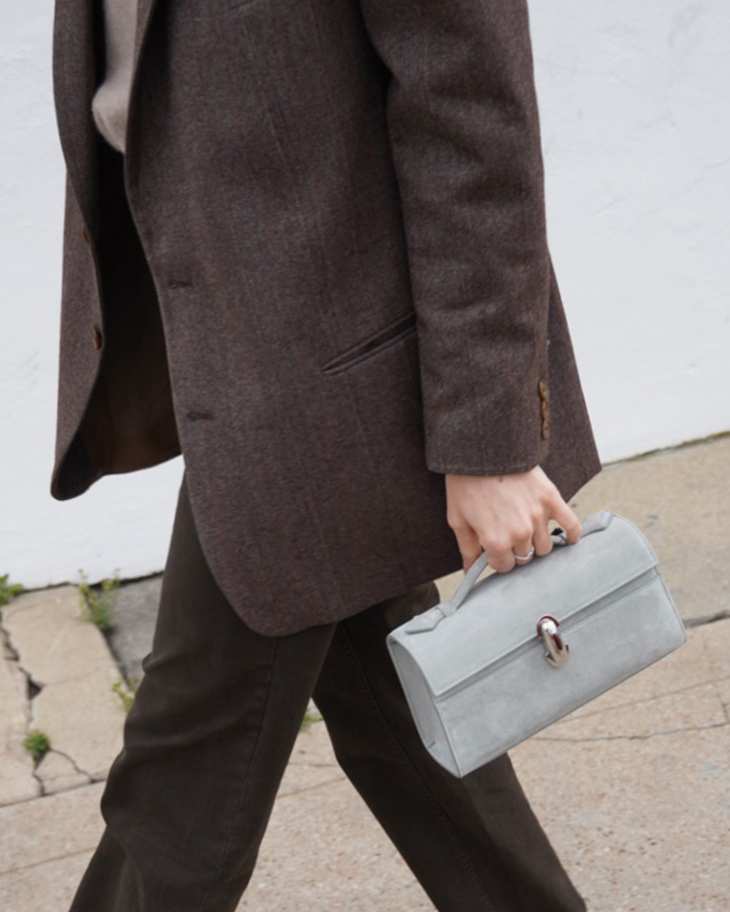 Close-up of tailored brown blazer with dark trousers and suede loafers%2C carrying a light gray suede top-handle bag on a sidewalk