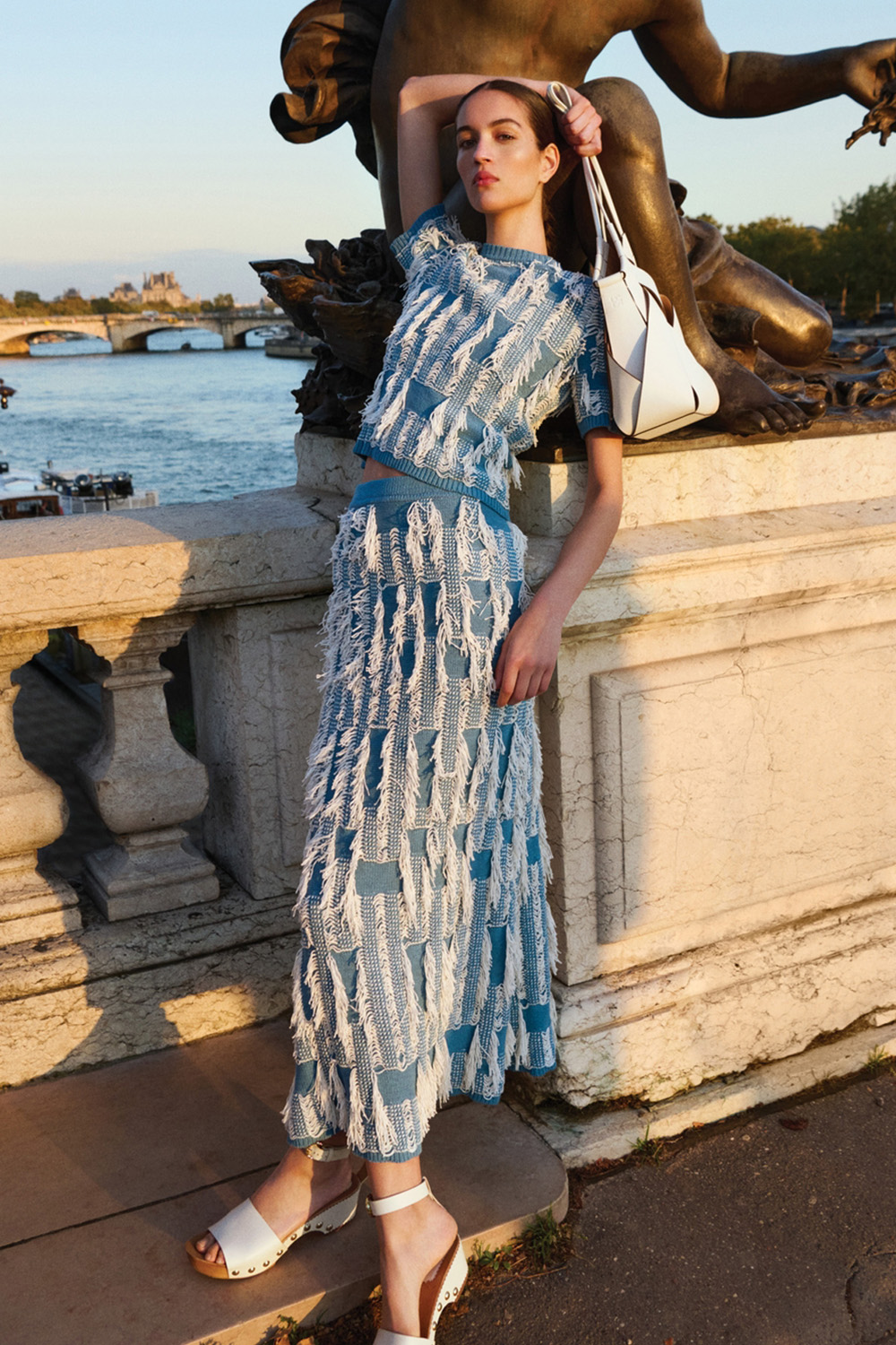 Ulla Johnson Spring 2025 collection features a model in a textured blue two-piece set%2C styled with a chic white handbag and platform sandals against a scenic riverside backdrop