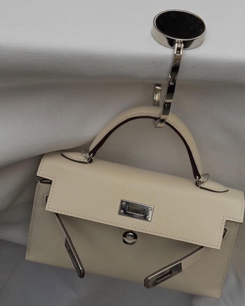 Cream structured top-handle bag with silver hardware and black charm%2C styled against soft white fabric%0A