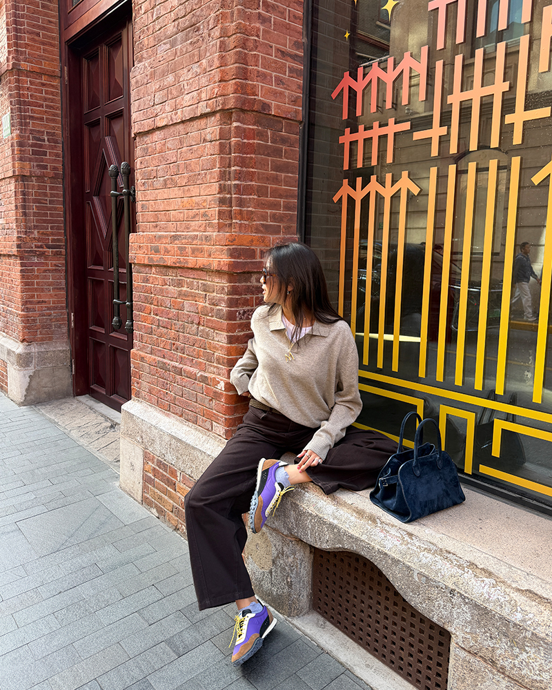 Woman in beige polo sweater with brown wide-leg trousers%2C colorful sneakers and suede tote%2C seated by brick storefront window