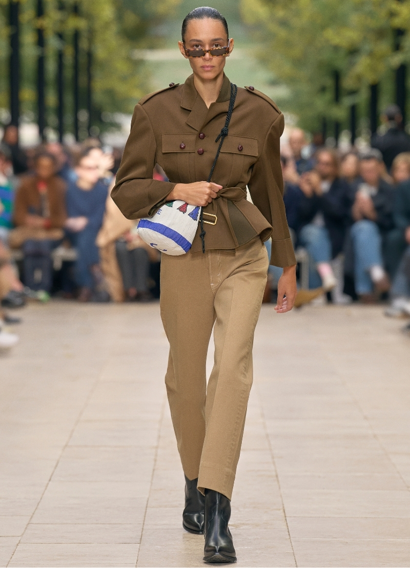 Model wears brown belted military jacket with tan trousers and ankle boots on an outdoor runway
