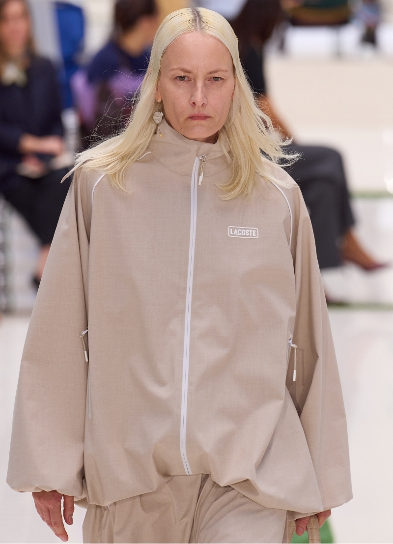 Model wearing beige Lacoste zip-up jacket and matching trousers, presented on a bright runway