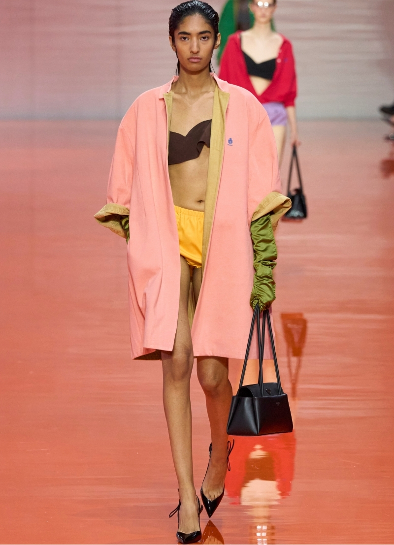 Model in coral oversized coat over bralette and shorts with black handbag, walking a glossy runway