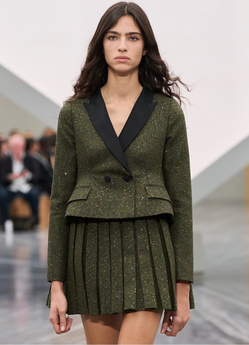 Runway model in green tweed blazer dress with black lapels and pleated skirt, fashion show
