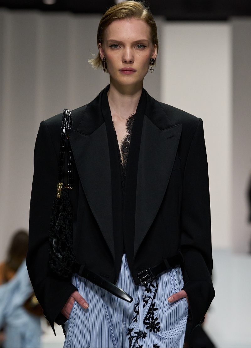 Runway model in black tuxedo blazer with lace top and striped trousers, carrying shoulder bag