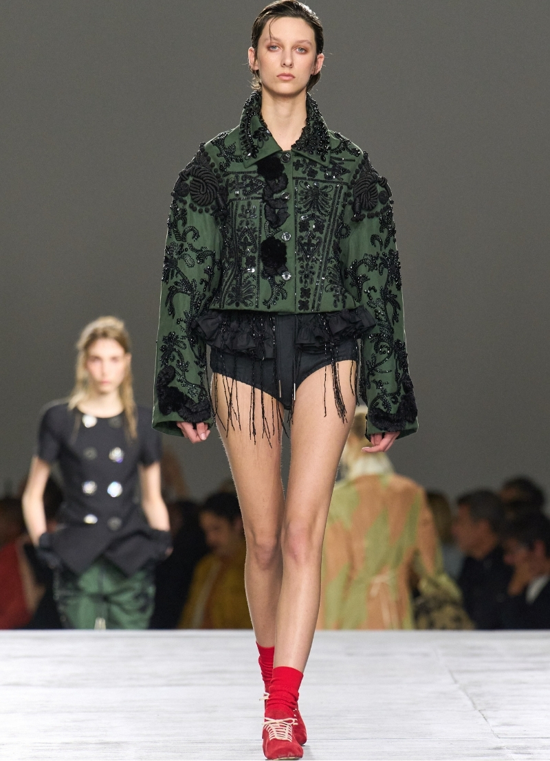 Runway model in embellished green cropped jacket with black fringe shorts, walking on runway