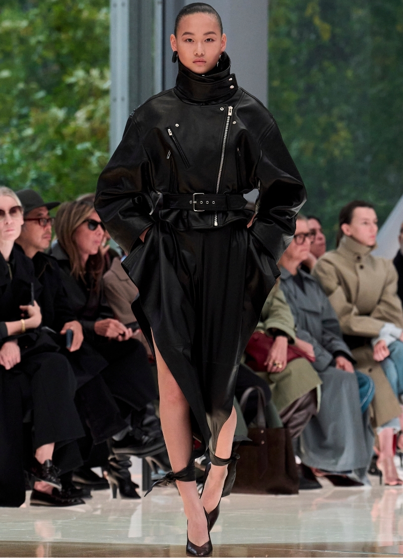 Model in black leather belted biker jacket dress with zip details and pointed heels on runway