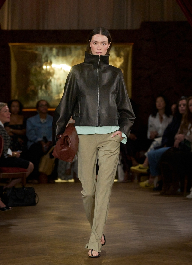 Model in cropped black leather jacket with beige trousers and clutch on indoor runway
