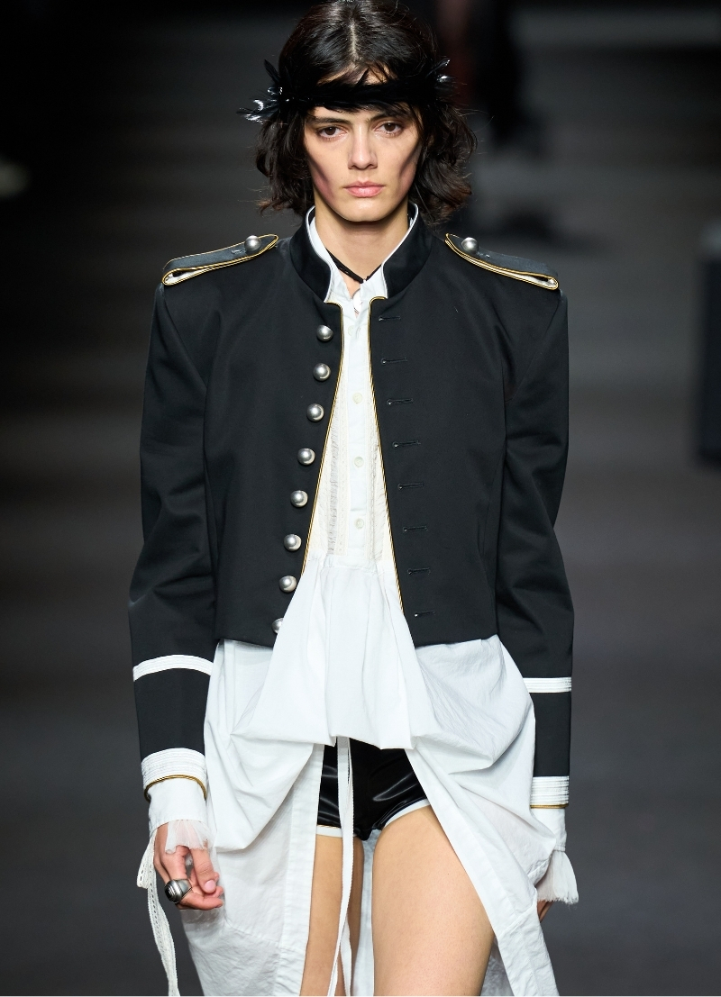 Model in black military jacket with silver buttons over white blouse and leather shorts on runway